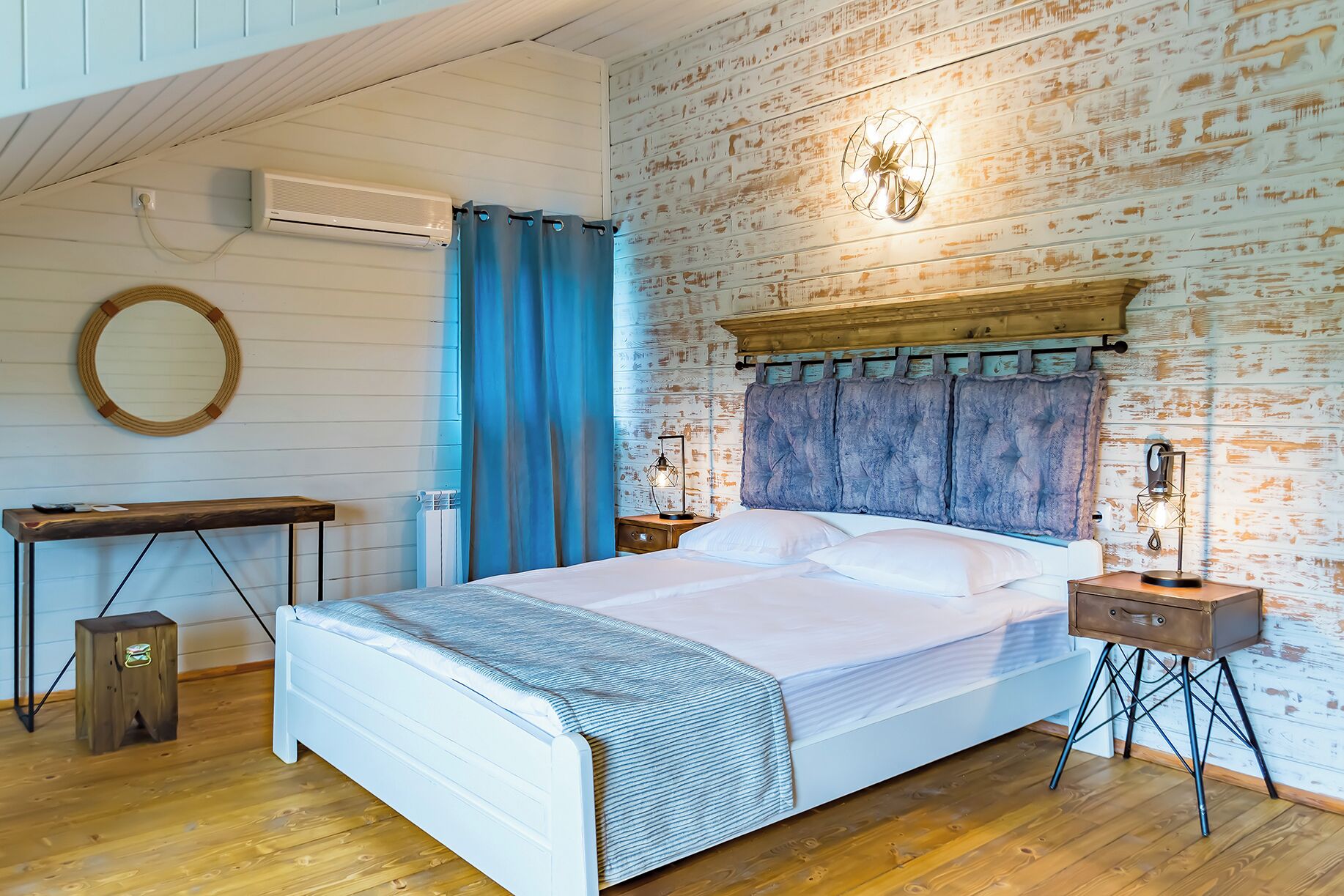 Quarto com cama, piso de madeira, espelho redondo, mesa de cabeceira, luminária e aquecedor fixado na parede.