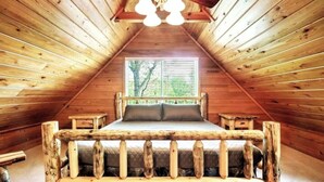 Interior de madeira com teto inclinado, cama com estrutura de madeira, janela com persianas e ventilador de teto.