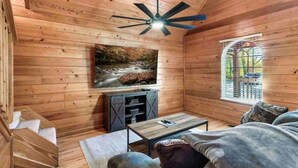 Interior acolhedor de uma cabana com paredes de madeira, ventoinha no teto, TV na parede e janela com vista para o verde, al ém de uma escada.