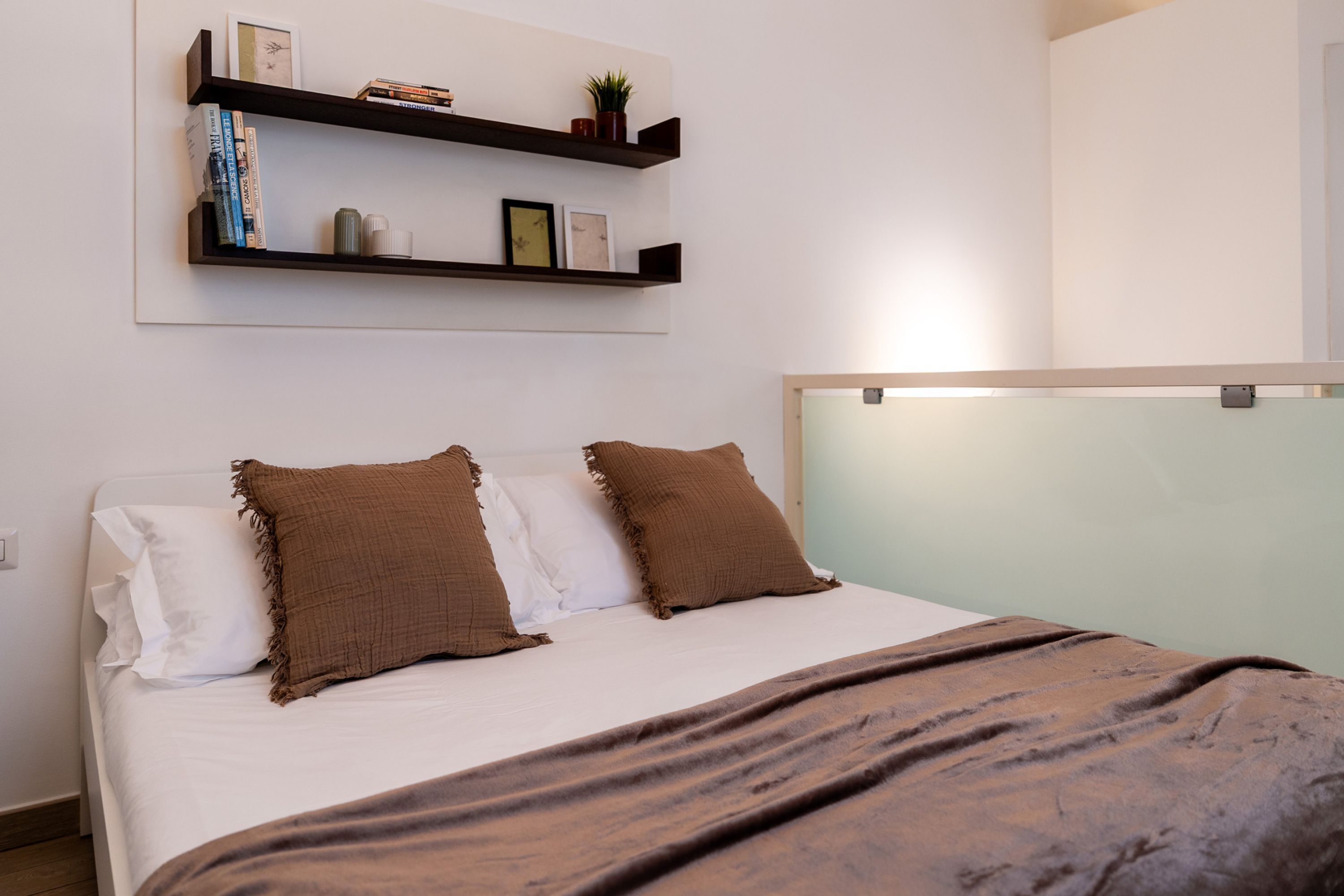 Cama bem arrumada com roupa de cama branca e castanha, mesa de cabeceira e prateleira com livros e objetos decorativos.