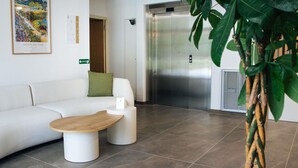 Uma área de recepção com sofá branco, mesa de centro de madeira, planta em vaso, elevador e uma obra de arte emoldurada na parede.