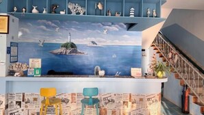 Uma área de recepção com um mural de um farol e mar, uma estante azul com objetos decorativos e um balcão com duas cadeiras.