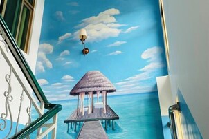 Uma escadaria com um mural representando uma construção de telhado de palha sobre pilotis na água, um corrimão e uma luminária fixada na parede.