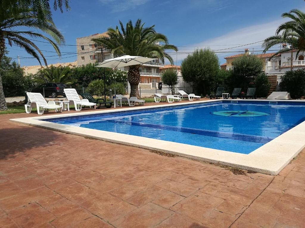 Uma piscina retangular com borda azulejada, rodeada por um pátio de tijolos. Há cadeiras de lounge brancas e uma palmeira ao fundo.