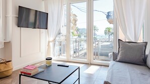 Uma sala de estar moderna com uma televisão de tela plana na parede, um sofá com almofadas, uma mesa de centro com livros e um controle remoto, e uma janela grande com cortinas.