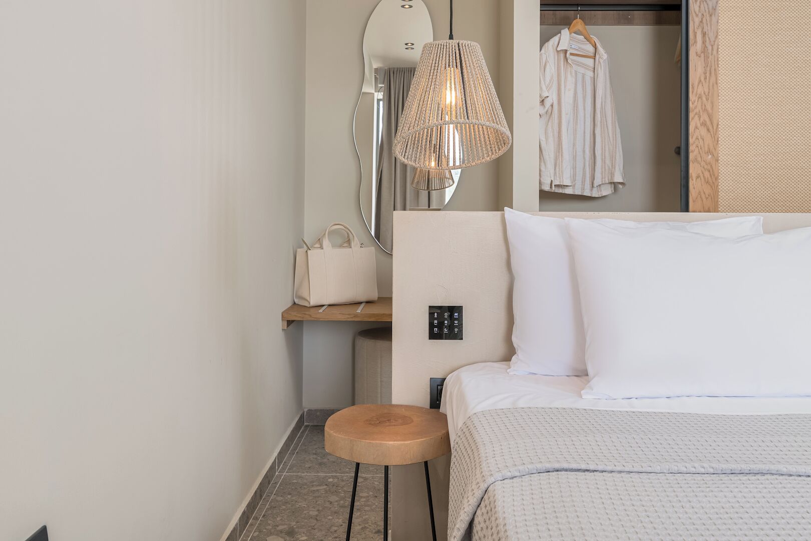 Quarto de hotel com cama, mesa de cabeceira, luminária pendente, espelho, armário e um banquinho pequeno.