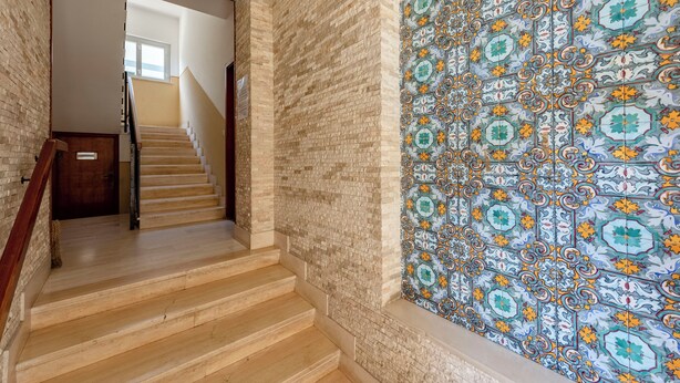 Uma escadaria com degraus de madeira e corrimão, que leva a um quarto com parede de tijolos e parede decorada com azulejos.
