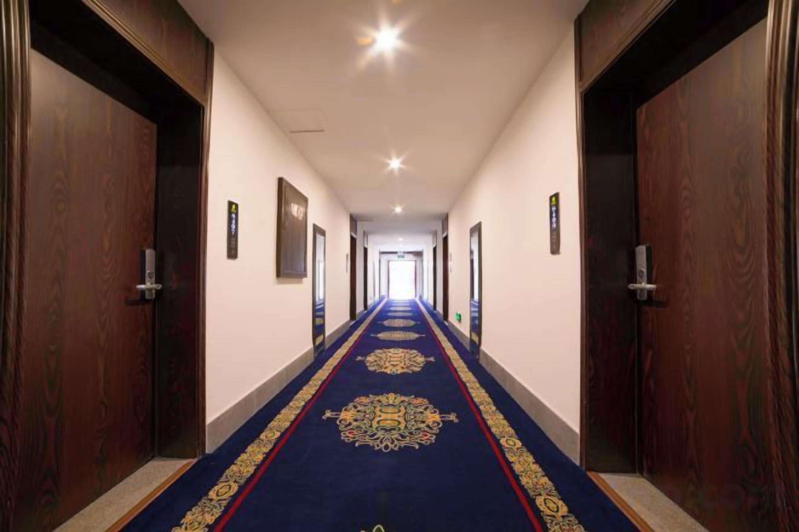 Corredor de hotel com carpete azul, portas de madeira e luminária no teto.