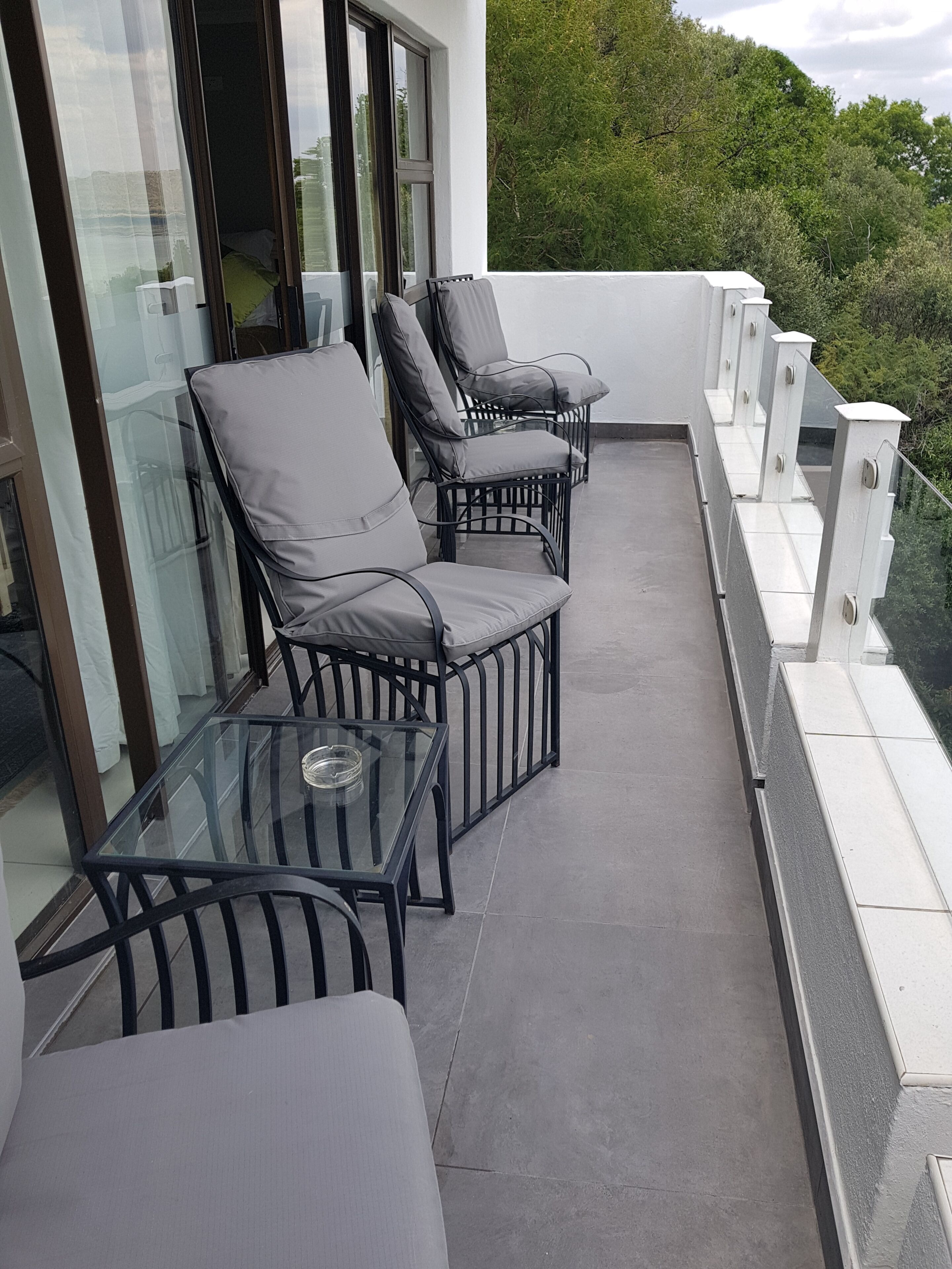 Varanda com mobiliário externo, grades de vidro e vista para o verde.