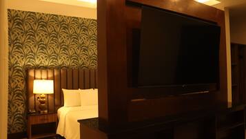 Quarto de hotel com uma televisão de tela plana grande fixada na parede, mesa de cabeceira com abajur, cama com roupa de cama branca e papel de parede estampado.