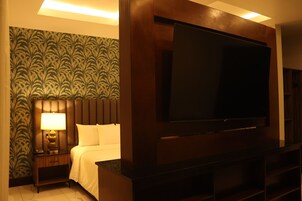 Quarto de hotel com uma televisão de tela plana grande fixada na parede, mesa de cabeceira com abajur, cama com roupa de cama branca e papel de parede estampado.