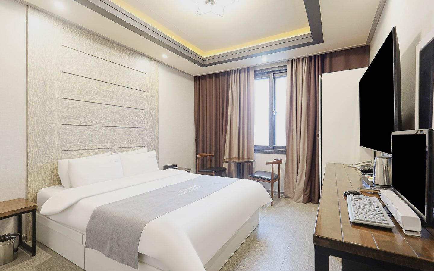Quarto de hotel com cama, escrivaninha com computador, cadeira, janela com cortinas e televisão.