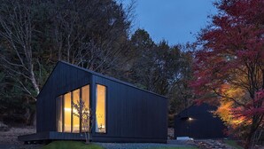 Uma cabana moderna, de cor escura, com grandes janelas de vidro, rodeada por árvores e um relvado bem cuidado.
