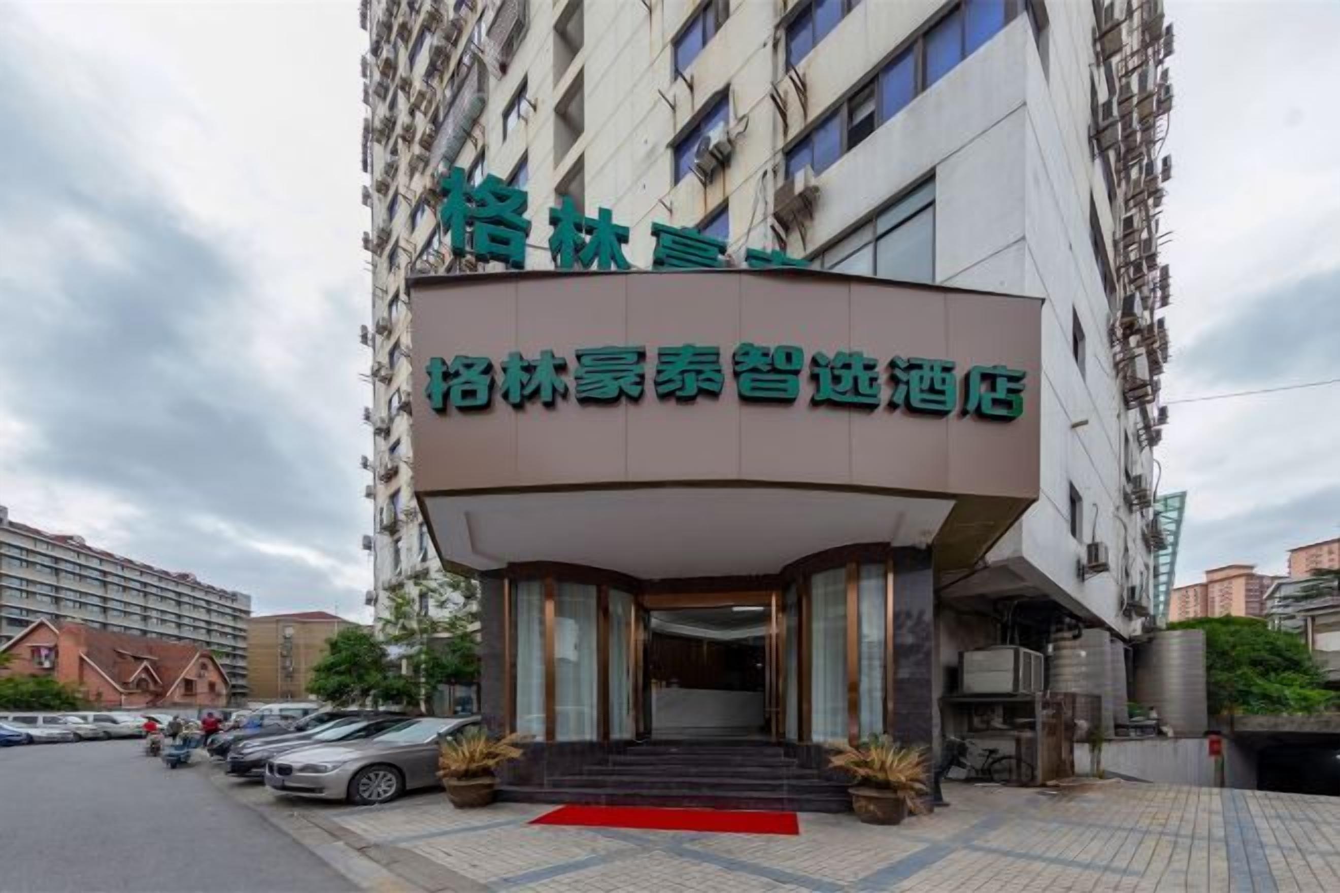 Entrada de hotel com fachada curva, tapete vermelho e estacionamento com carros.