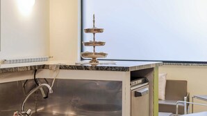 Cozinha moderna com pia de aço inoxidável, janela com persianas e pequena área de jantar com cadeiras.