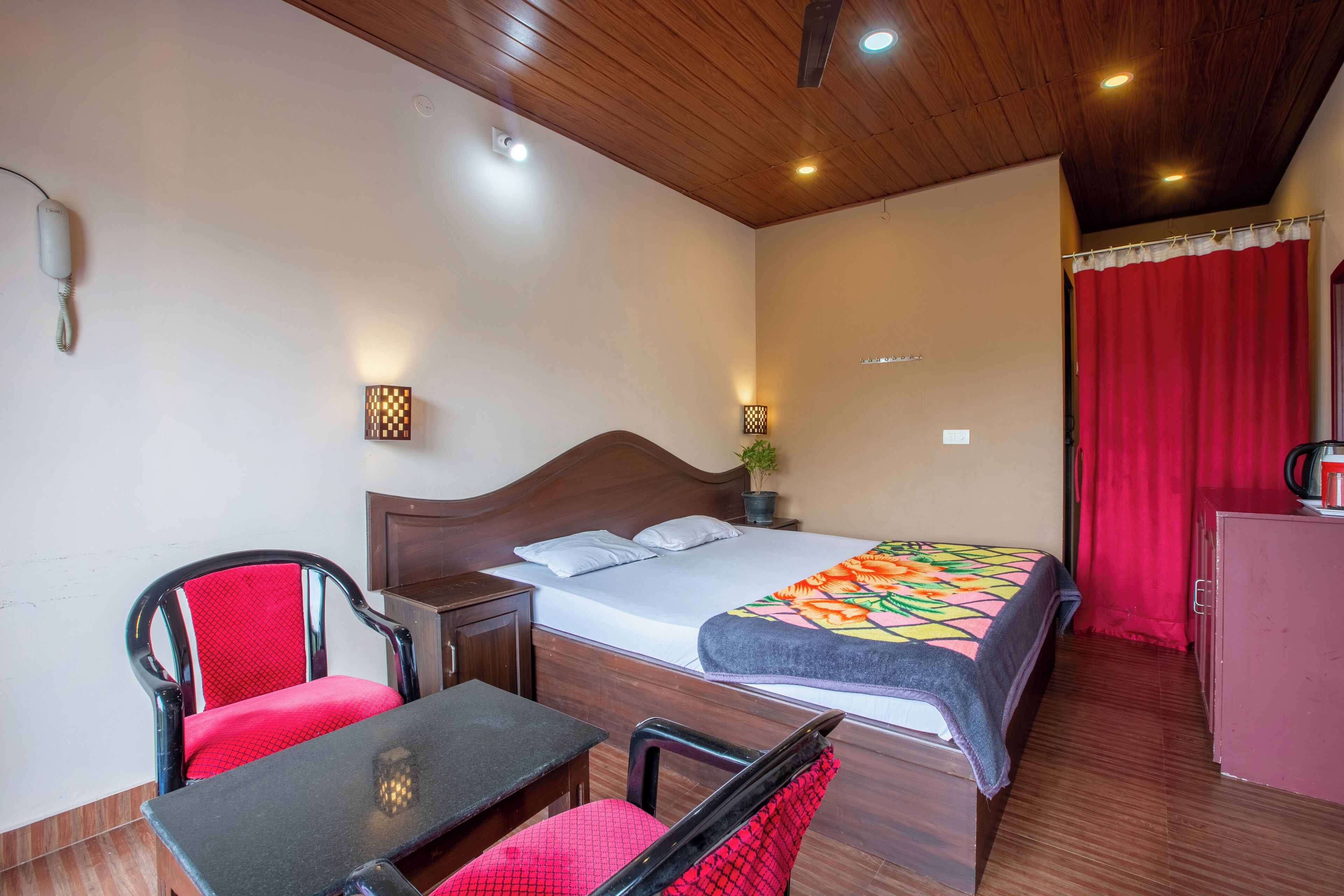 Um quarto com teto de madeira, uma cama com manta colorida, uma escrivaninha com cadeira, uma mesinha de cabeceira com abajur e uma cortina vermelha.