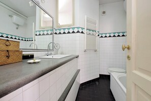 Casa de banho com lavatório, espelho e azulejos brancos com borda geométrica.