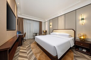 Quarto de hotel com uma cama grande, escrivaninha, cadeira, televisão e mesa de cabeceira com abajur.