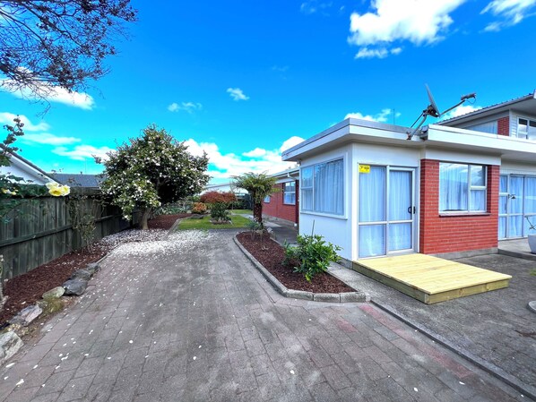 Uma área residencial com calçada pavimentada, cerca de madeira e um pequeno jardim com plantas floridas.