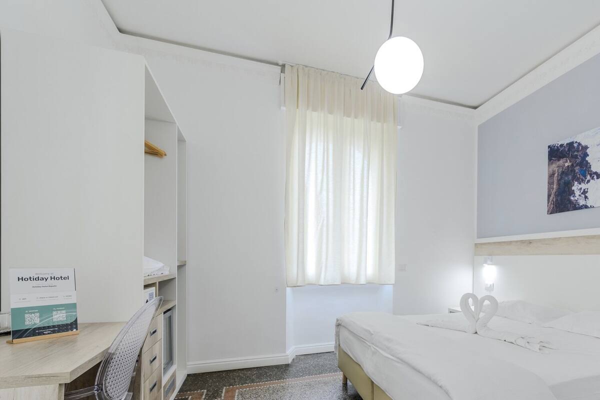 Quarto de hotel com cama, escrivaninha, cadeira, janela com cortinas, uma obra de arte na parede e uma placa sobre a escrivaninha.
