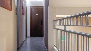 Um corredor com uma porta de madeira no final, um corrimão de metal à direita e um teto com iluminação embutida.