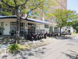 Um edifício com uma fileira de motocicletas estacionadas, uma árvore em primeiro plano e um passeio pavimentado.