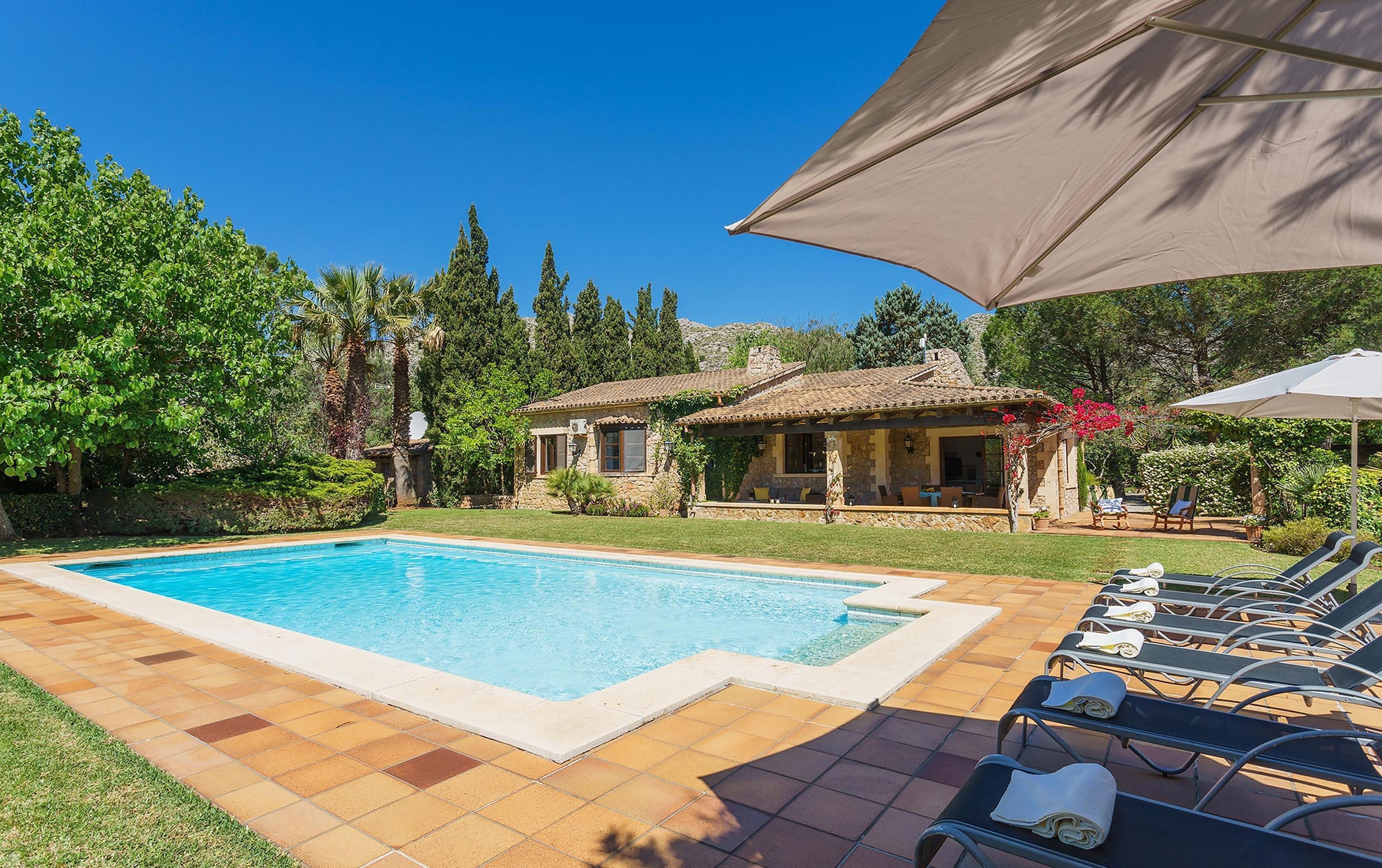 Uma piscina com espreguiçadeiras e guarda-sóis, cercada por vegetação e edifícios de pedra.