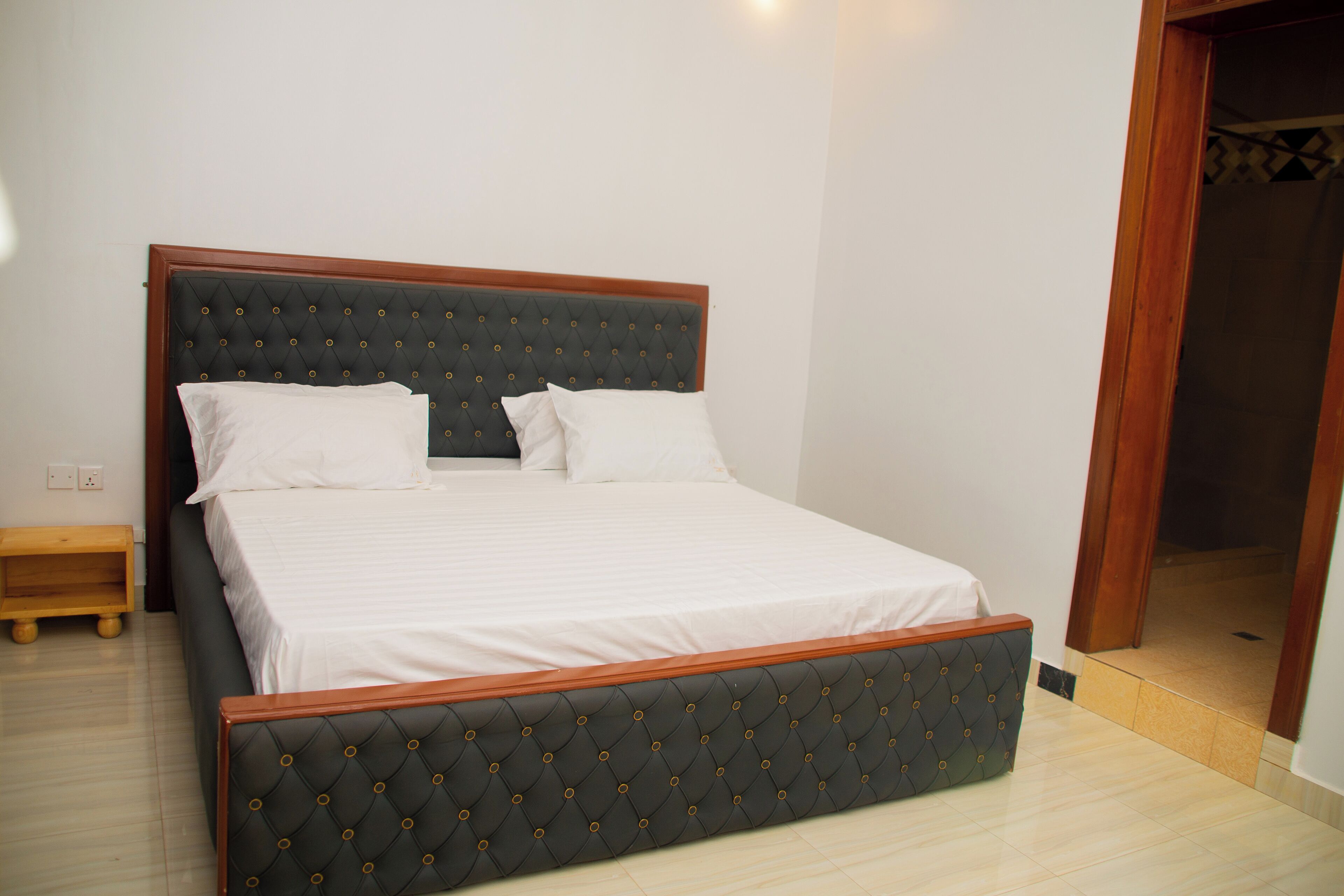 Cama bem arrumada com cabeceira escura e roupa de cama branca em quarto com piso de madeira clara e mesa de cabeceira de madeira.