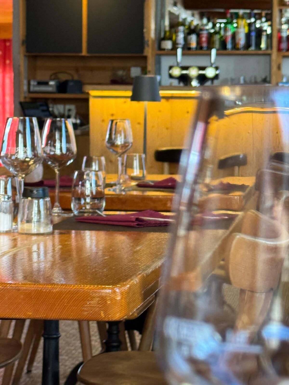 Interior de restaurante com mesas e cadeiras de madeira, copos de vinho e um bar ao fundo.
