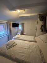 Quarto de hotel com duas camas, televisão fixada na parede, janela com cortinas e porta com uma toalha pendurada.