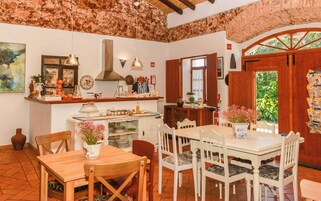 Uma zona de refeições rústica, com uma mesa e cadeiras de madeira, uma bancada de cozinha com diversos objetos e vista para o verde através de uma porta de madeira.
