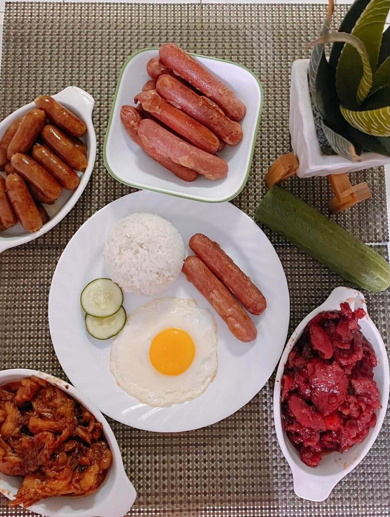 Uma refeição com salsichas, arroz, um ovo frito, fatias de pepino e uma tigela de molho vermelho.
