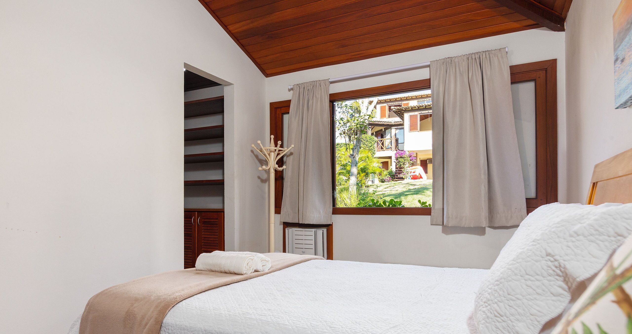 Quarto com cama, janela com cortinas, um cabide e vista para um edifício.