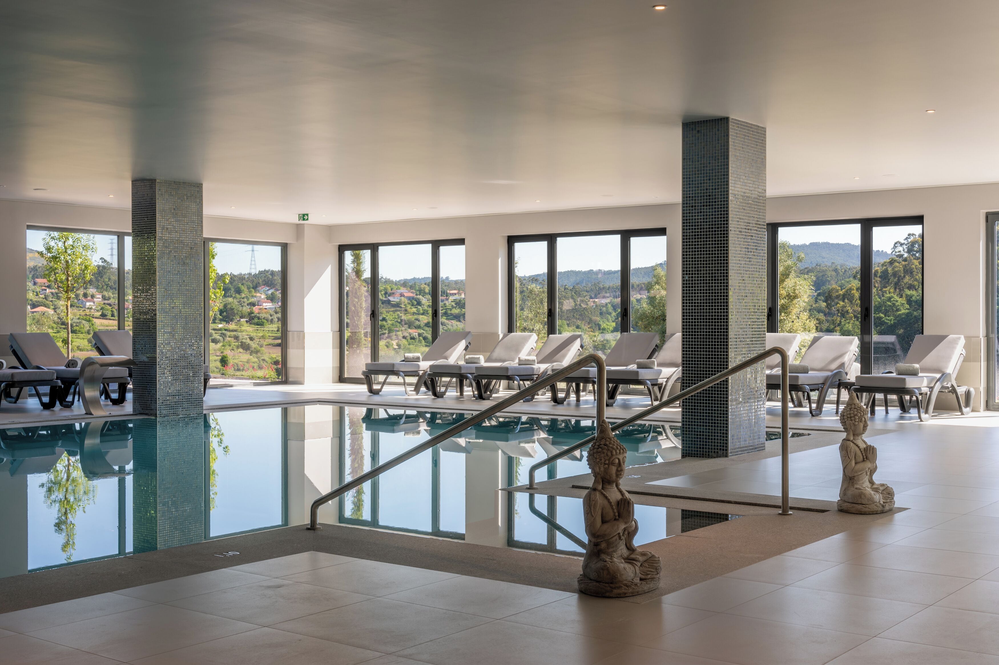 Área moderna de piscina interna com vista para a paisagem circundante, cadeiras de descanso e uma estátua de Buda.