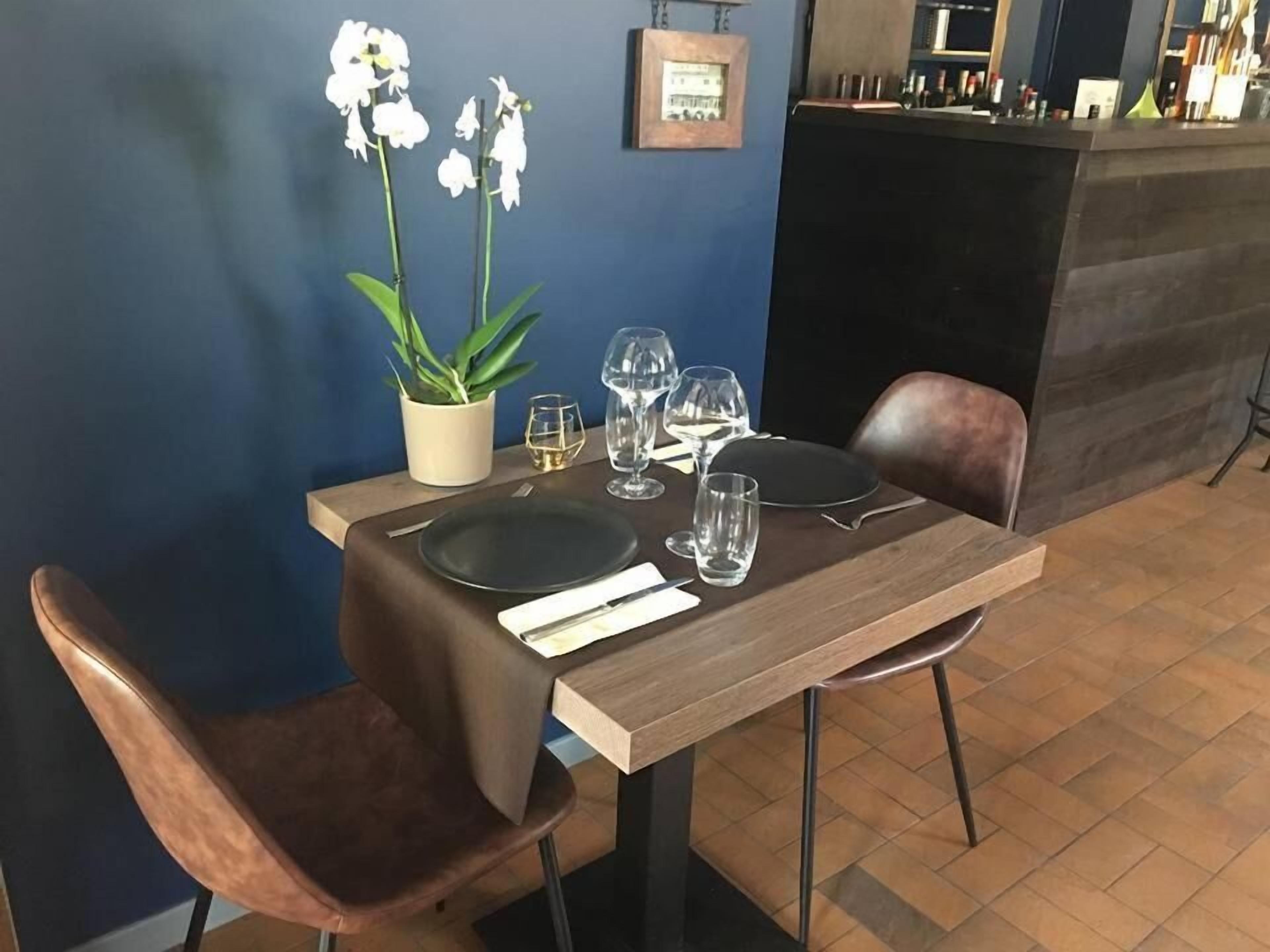Mesa de jantar posta com pratos, talheres e copos de vinho, com uma planta em vaso e um quadro na parede.
