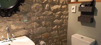 Casa de banho com parede de pedra, uma pia, sanita e prateleira com artigos de higiene pessoal.