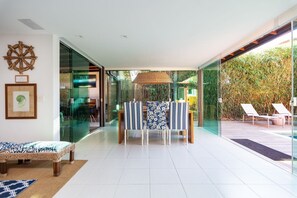 Interior moderno com divisória de vidro, área de refeições com cadeiras listradas e vista para uma piscina e vegetação lá fora.