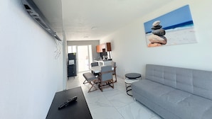 Uma sala de estar moderna com um sofá cinzento, uma mesinha e um comando de televisor numa bandeja preta. Há um quadro na parede com uma pintura de pedras empilhadas.