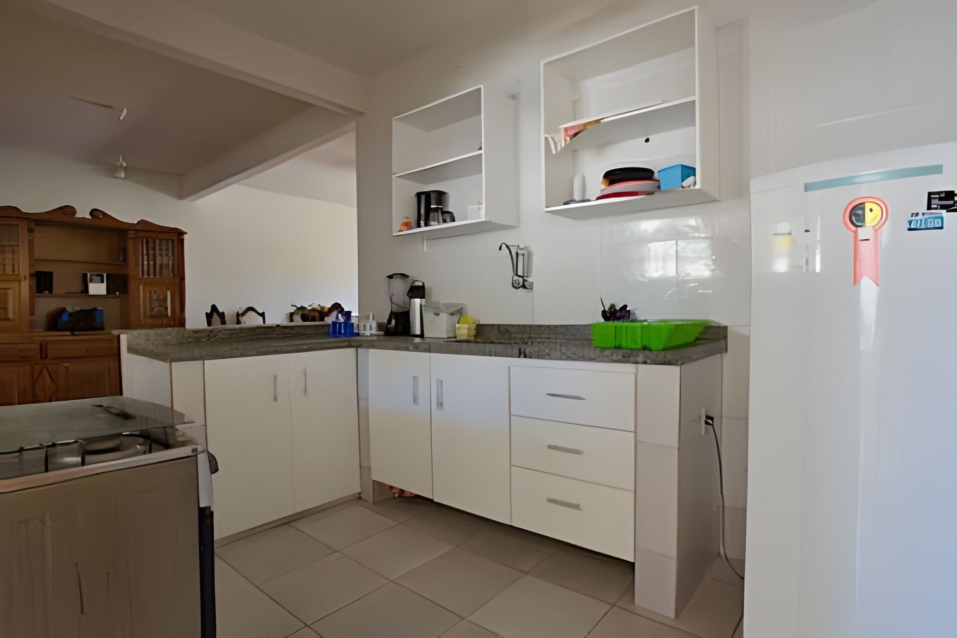 Cozinha com armários brancos, bancada e geladeira. Há prateleiras acima da bancada e um armário de madeira à esquerda.