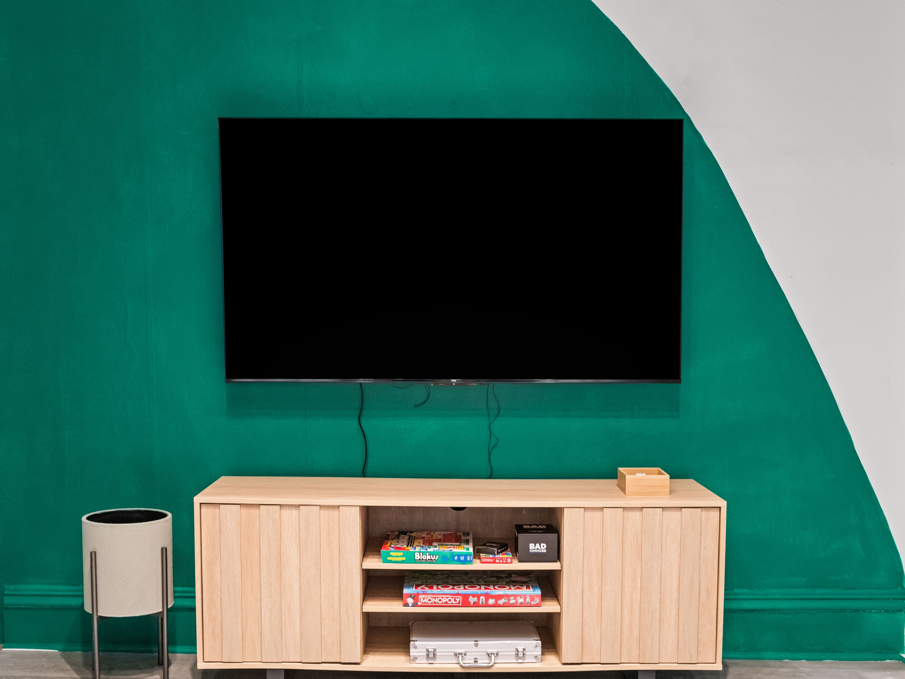 Quarto com televisão de tela grande na parede verde, um armário de madeira com jogos de tabuleiro e uma luminária cilíndrica.