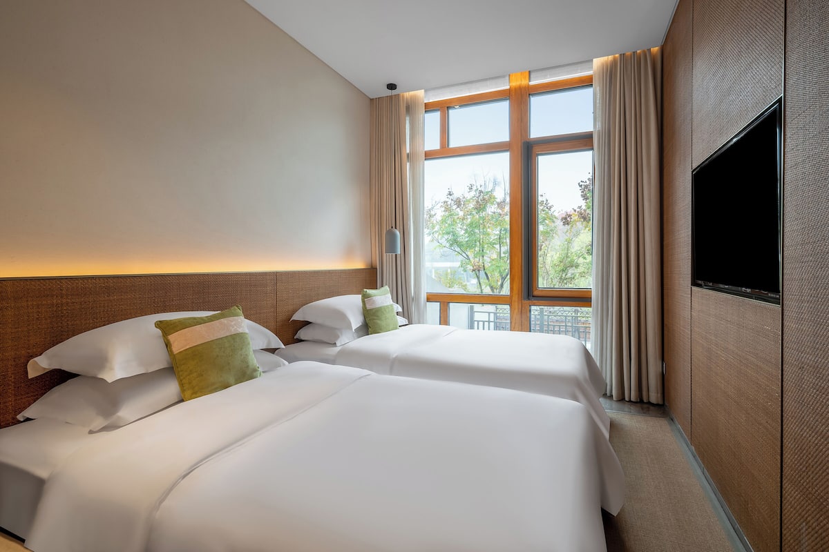 Quarto de hotel com duas camas, uma janela grande com cortinas, televisão de tela plana e vista para área verde.