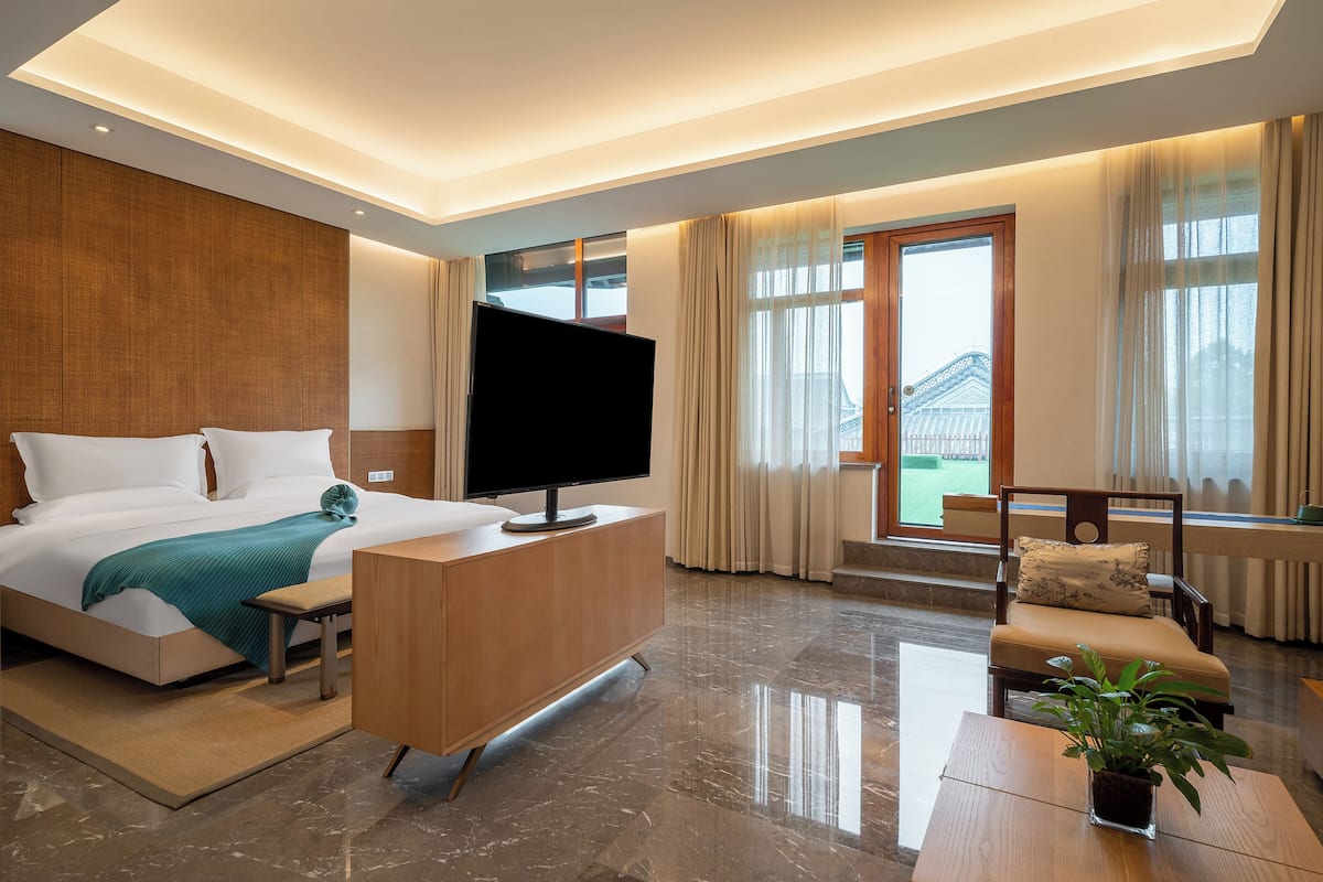 Quarto de hotel moderno com uma cama grande, televisão de tela plana, escrivaninha de madeira, poltrona confortável e vista para um edifício.