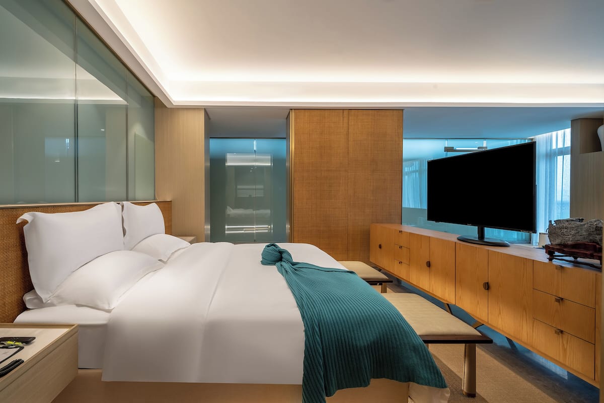 Quarto de hotel moderno com uma cama grande, uma televisão, uma cômoda de madeira e um banco.