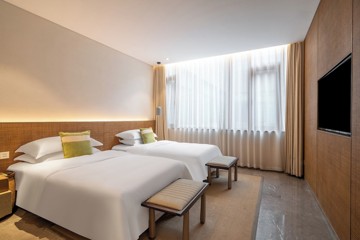 Quarto de hotel com duas camas, televisão, mesa de cabeceira e um banco.
