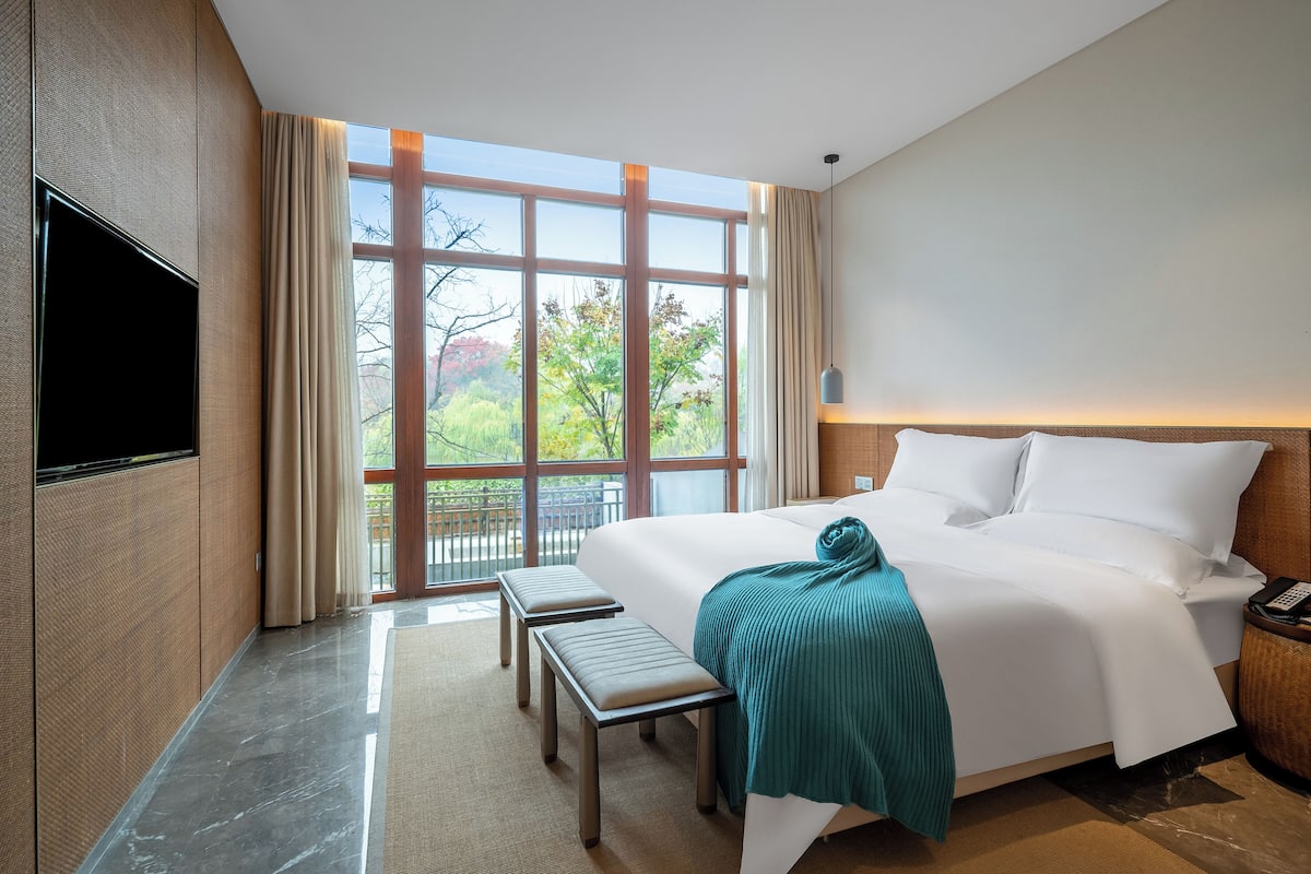 Quarto de hotel com uma janela grande, uma cama, uma TV e um banquinho.