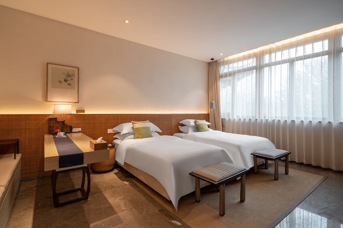 Quarto de hotel com duas camas, uma mesa com cadeira, uma mesinha e uma janela com cortinas.