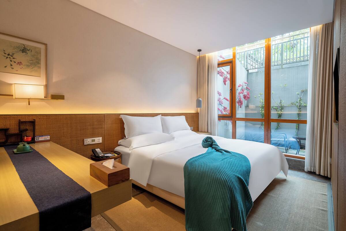 Quarto de hotel com cama, mesa de cabeceira, luminária e vista para o exterior.
