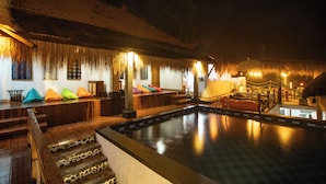 Uma área de piscina com degraus que levam à água, um telhado de palha e um espaço de bar com almofadas coloridas.