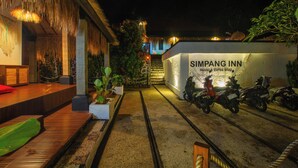 Cena noturna em Simpang Inn, com um edifício de telhado de palha, um Hostler & Coffee Shop e motocicletas estacionadas na frente.
