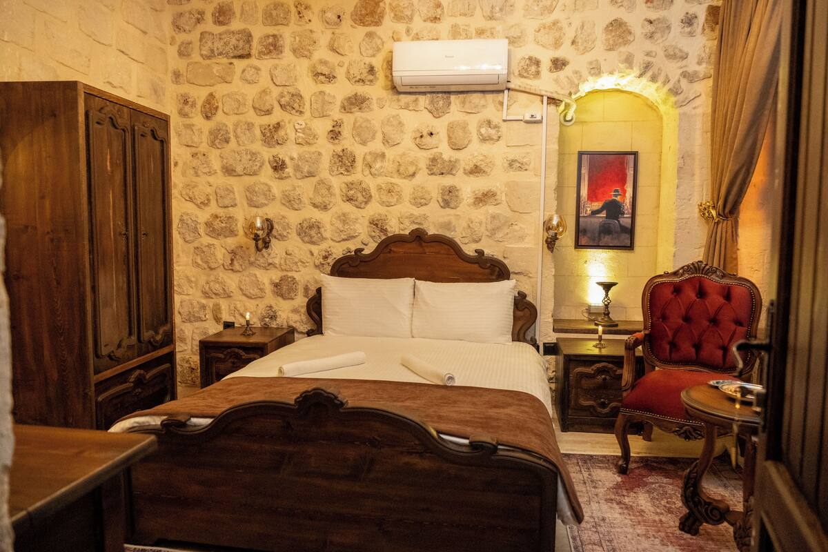 Quarto com parede de pedra, cama de madeira, uma cadeira vermelha e uma mesa de cabeceira com abajur.